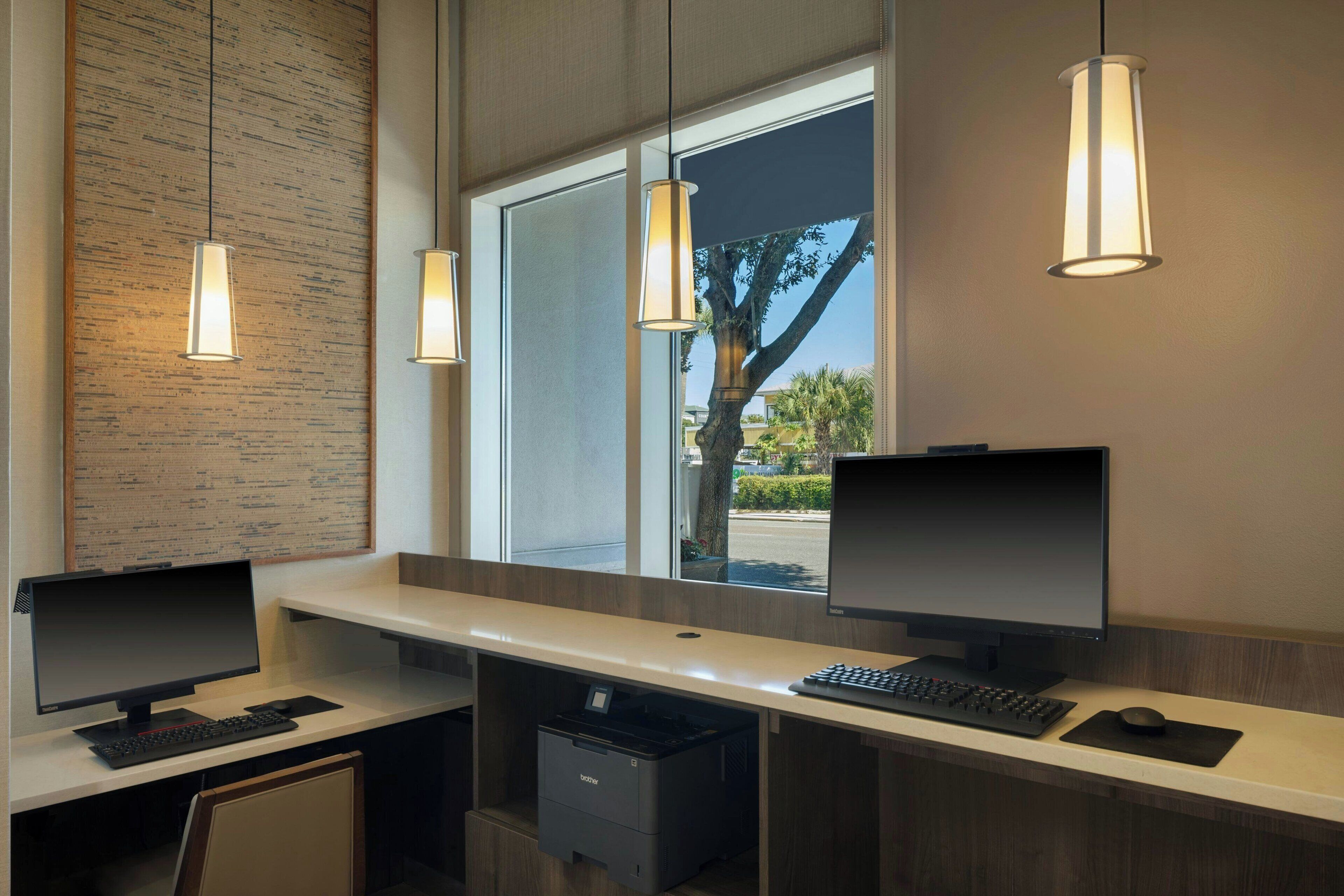 Espaço de escritório moderno com duas mesas, cada uma equipada com monitor, teclado e rato. Duas luminárias pendentes iluminam as mesas, e uma grande janela oferece vista para o exterior.