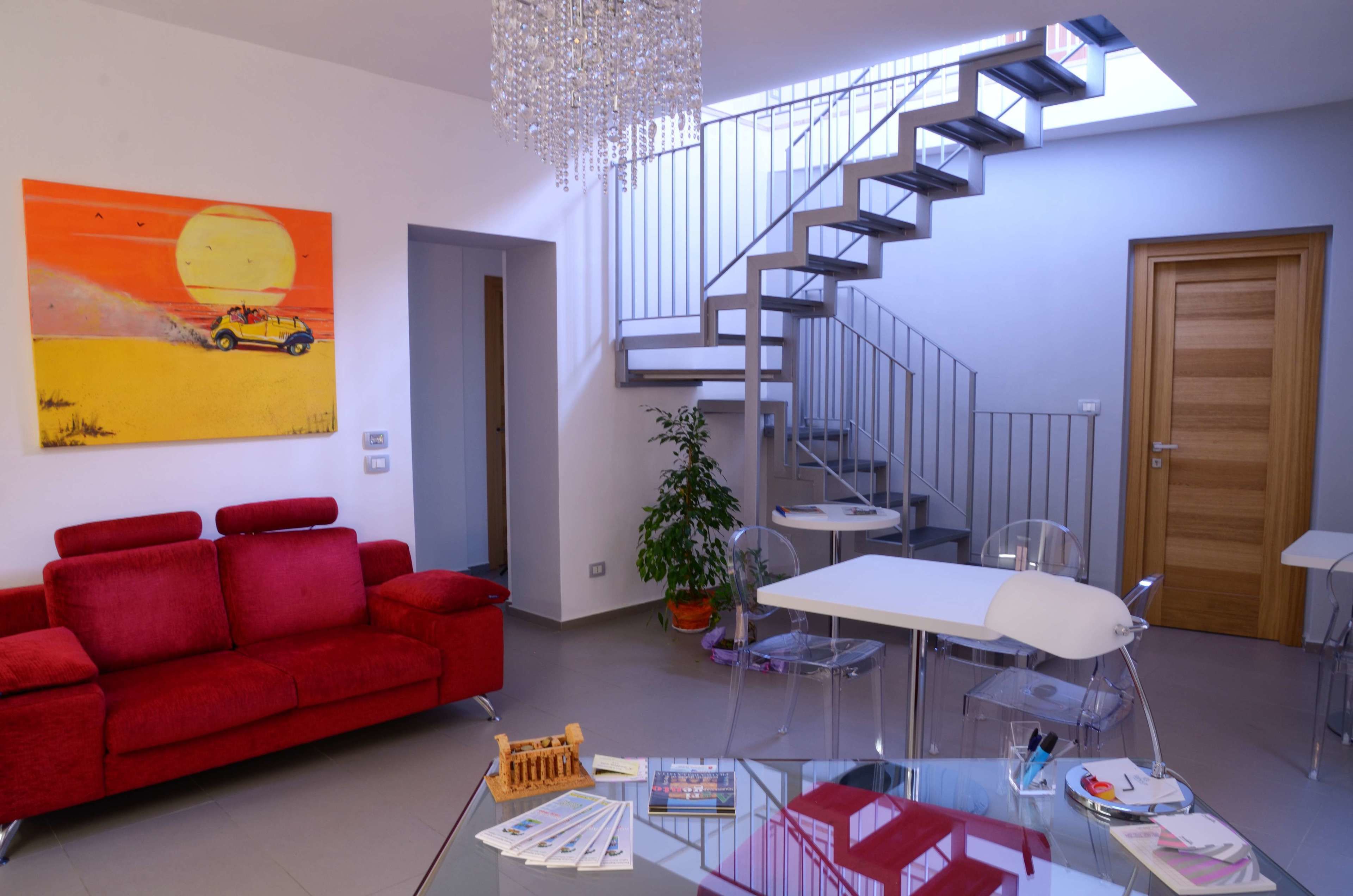 Uma sala de estar moderna com um sofá vermelho, uma mesa de centro de vidro, uma mesa branca e uma escada que leva a um andar superior.