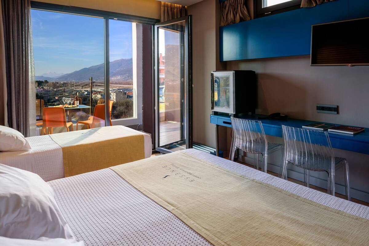 Quarto de hotel com uma cama grande, uma mesa com computador, televisão e vista para a cidade através de uma porta de correr.