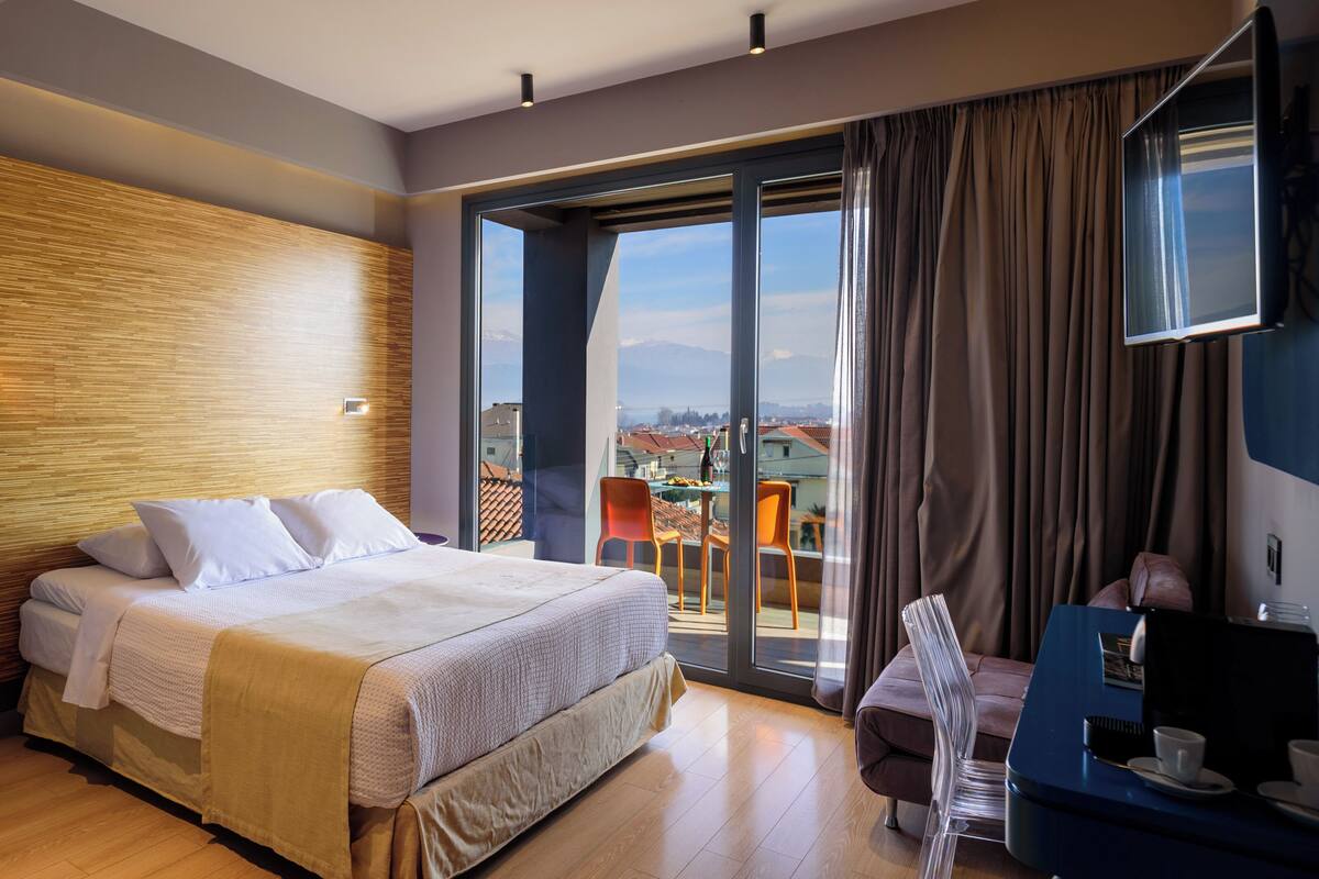 Quarto de hotel com uma cama grande, varanda com vista para a cidade, escrivaninha com cafeteira e televisão na parede.