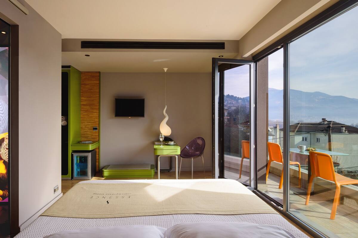 Quarto de hotel moderno com uma cama grande, uma televisão de tela plana, uma mesinha com abajur e uma varanda com cadeiras.