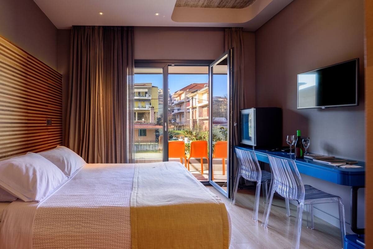 Quarto de hotel com cama, escrivaninha com computador, televisão e varanda com cadeiras laranjas.