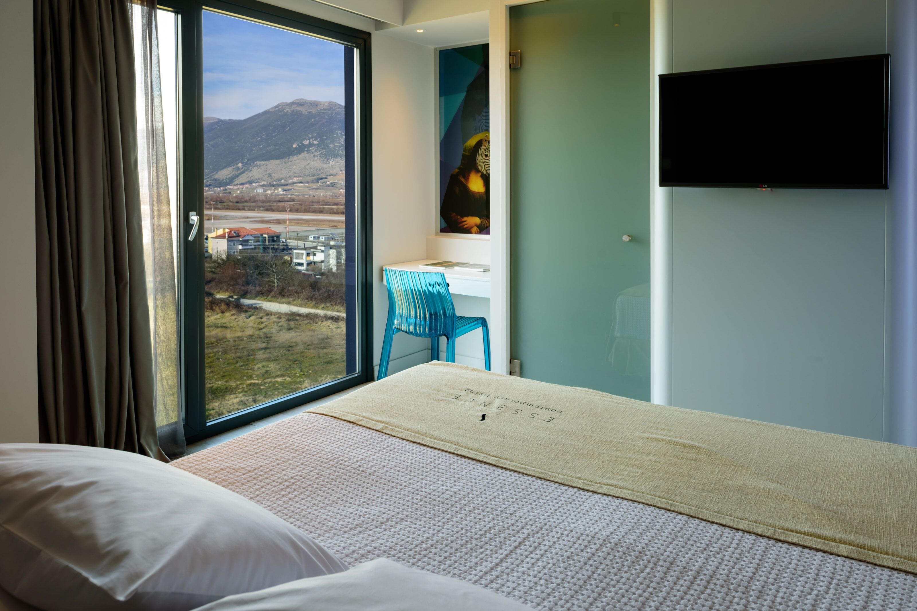 Quarto de hotel com cama, janela com cortinas, escrivaninha com cadeira, um quadro e uma televisão.