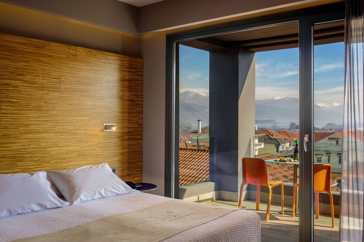 Quarto de hotel moderno com uma cama grande, uma parede de destaque em madeira e uma varanda com vista para a cidade e montanhas.