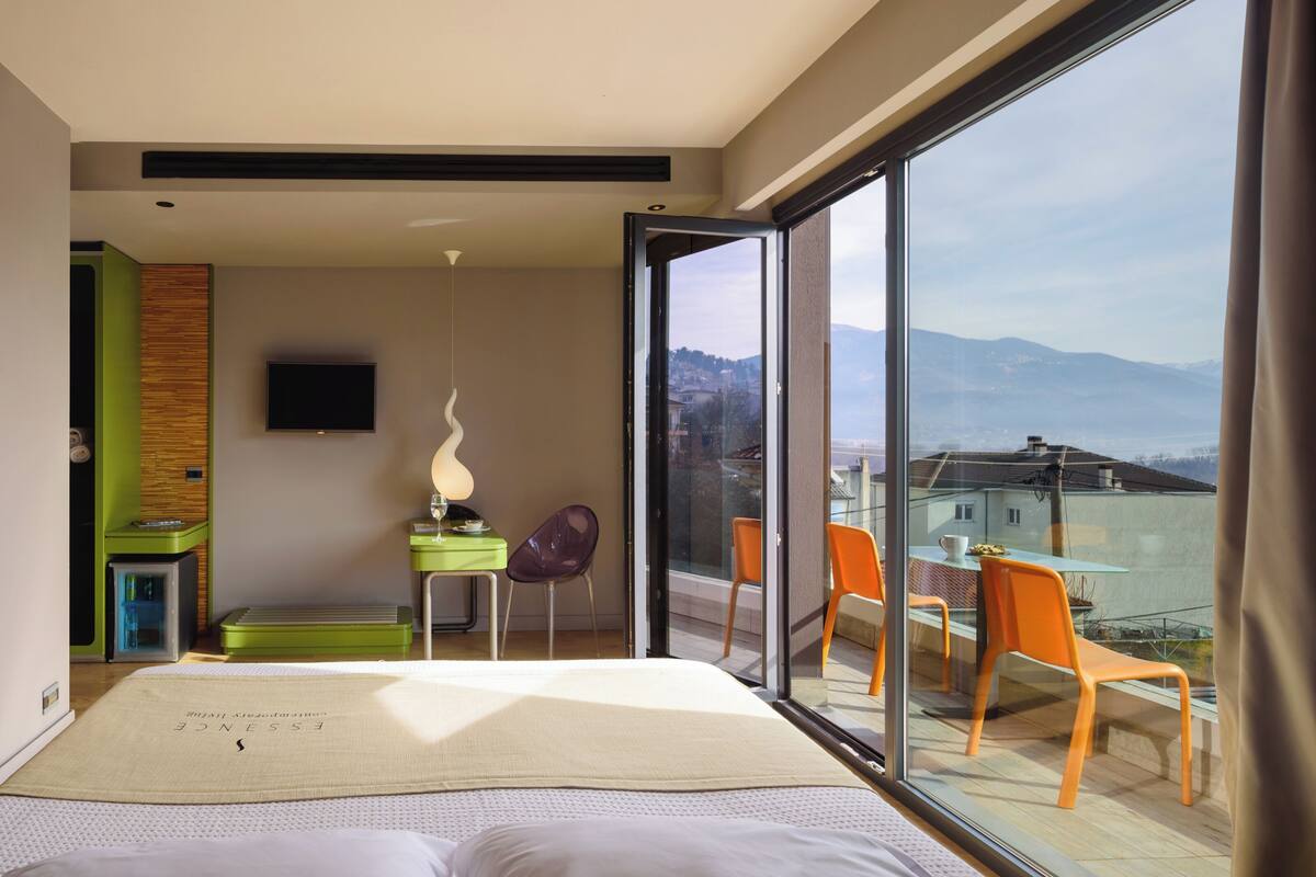 Quarto de hotel moderno com uma cama grande, televisão de tela plana, uma mesinha com cadeira e uma varanda com cadeiras laranjas.
