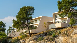 Um edifício moderno com várias varandas e telhado inclinado, rodeado por árvores e terreno rochoso.