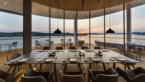 Uma sala de jantar com mesas preparadas para uma refeição, grandes janelas com vista para o pôr do sol sobre a água, e iluminação pendente.