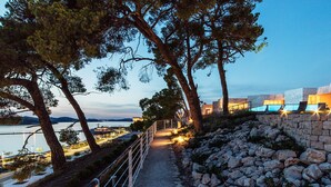 Uma passarela costeira com muros de pedra e árvores, que conduz a um edifício moderno com grandes janelas e iluminação externa.