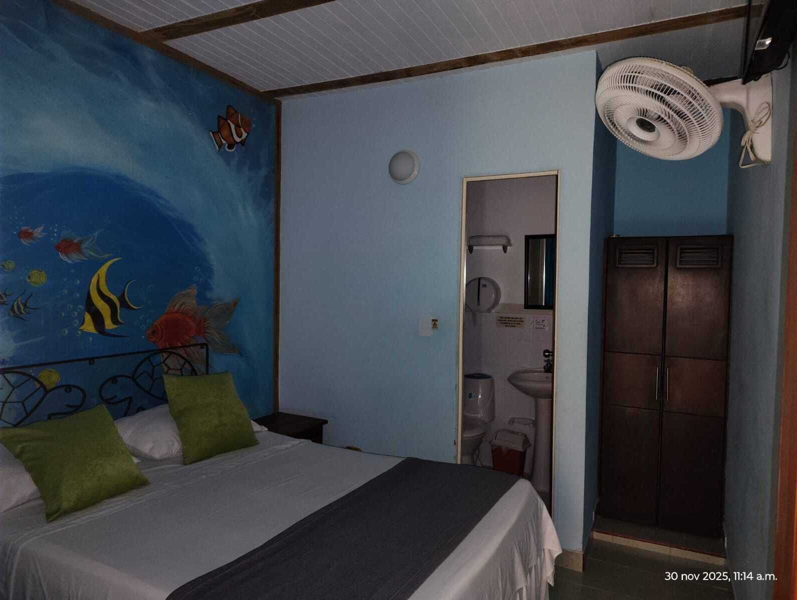 Quarto com cama, almofadas verdes, parede azul com mural de peixes, ventilador, guarda-roupa e banheiro visível por uma porta aberta.