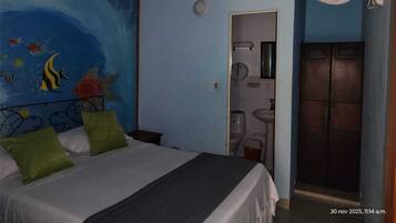 Quarto com cama, almofadas verdes, parede azul com mural de peixes, ventilador, guarda-roupa e banheiro visível por uma porta aberta.