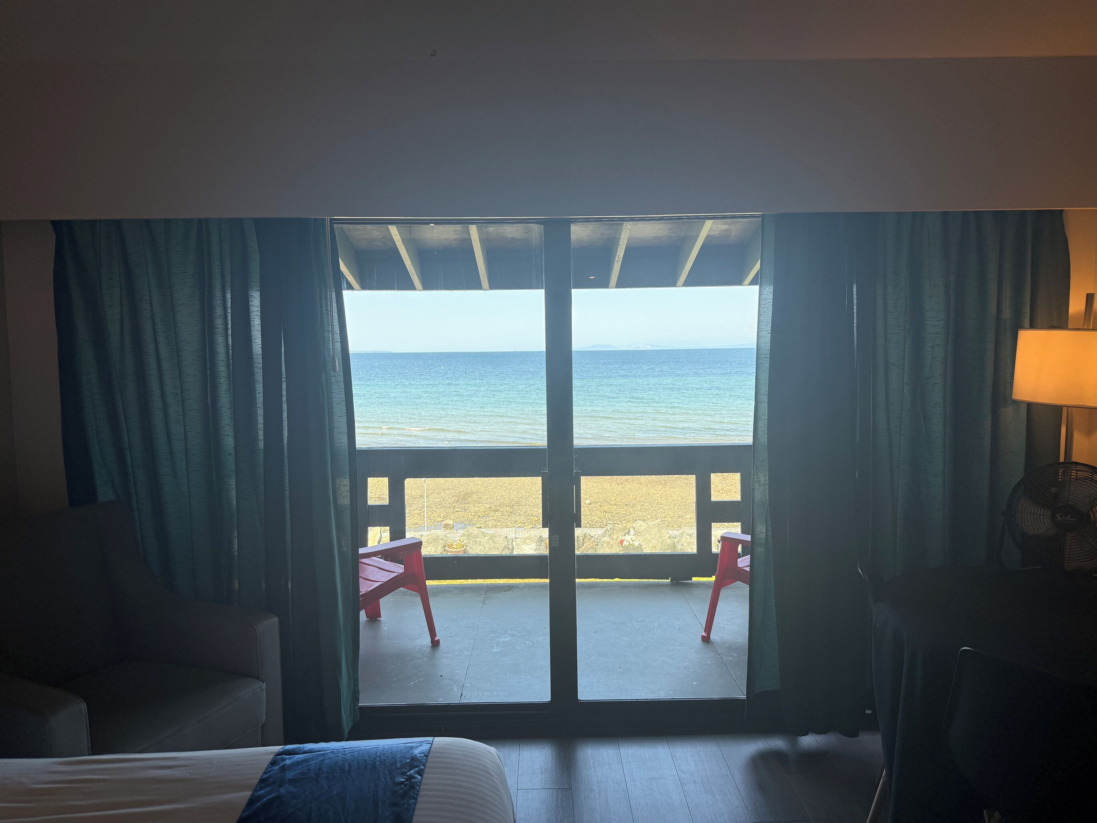 Quarto com cama, escrivaninha e vista para a praia através de uma porta de vidro.