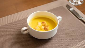 Uma tigela branca de sopa com croutons sobre um tapete texturizado, uma faca e um copo de água em uma mesa de madeira.