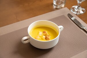Uma tigela branca de sopa com croutons sobre um tapete texturizado, uma faca e um copo de água em uma mesa de madeira.