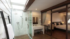 Quarto de hotel com chuveiro em gabinete de vidro, piso de madeira, cama com cabeceira, mesa de cabeceira e espelho.
