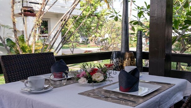 Mesa posta para dois, com toalha branca, copo de vinho tinto, xícara branca, prato com guardanapo e pequeno vaso de flores.