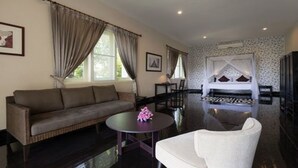 Uma sala de estar moderna com um sofá, uma mesa de centro redonda e uma poltrona branca. Uma cama com dossel pode ser vista ao fundo.