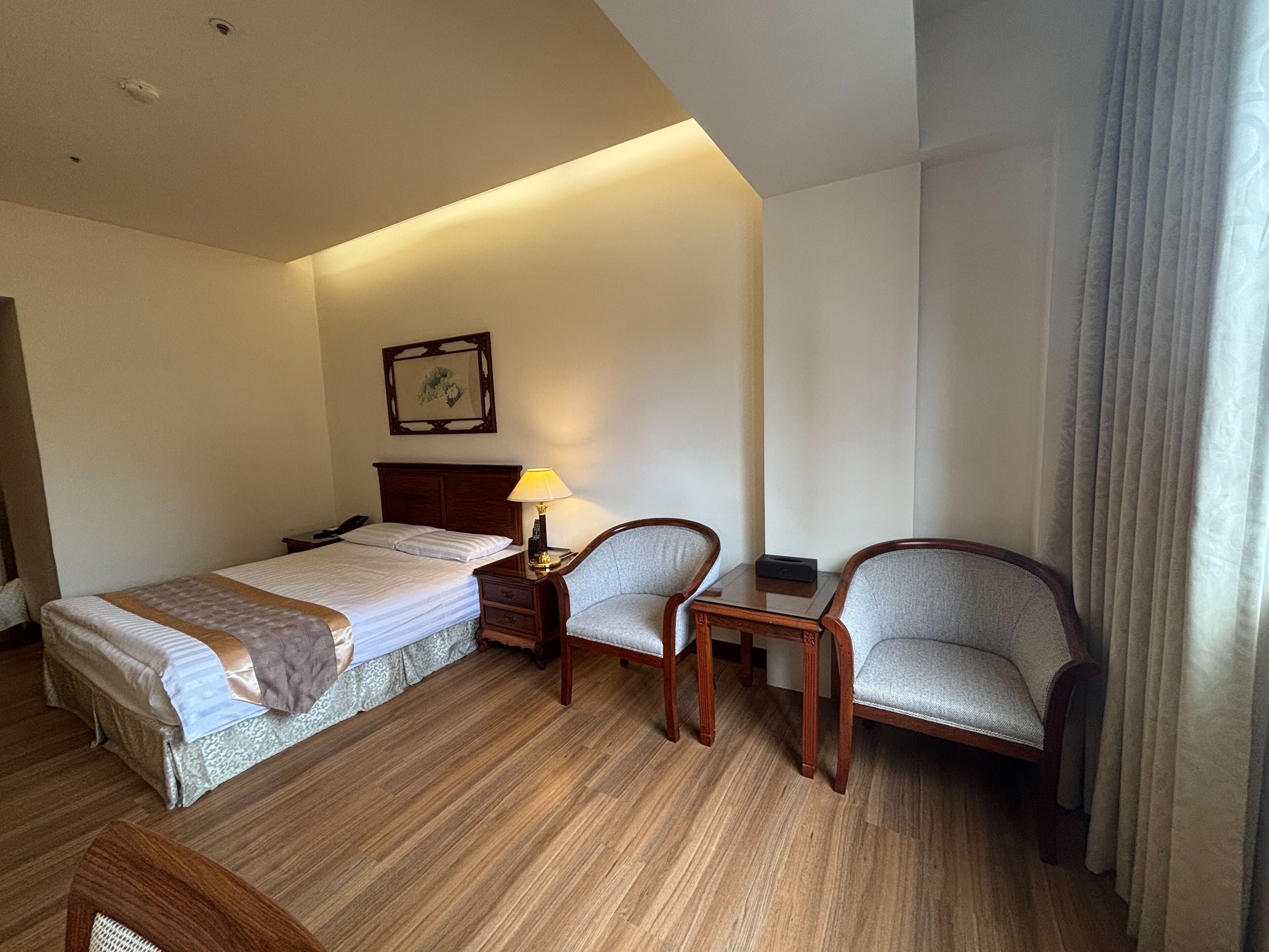 Quarto de hotel com cama, duas cadeiras, uma mesinha, abajur, mesa de cabeceira e um quadro na parede.