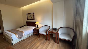 Quarto de hotel com cama, duas cadeiras, uma mesinha, abajur, mesa de cabeceira e um quadro na parede.