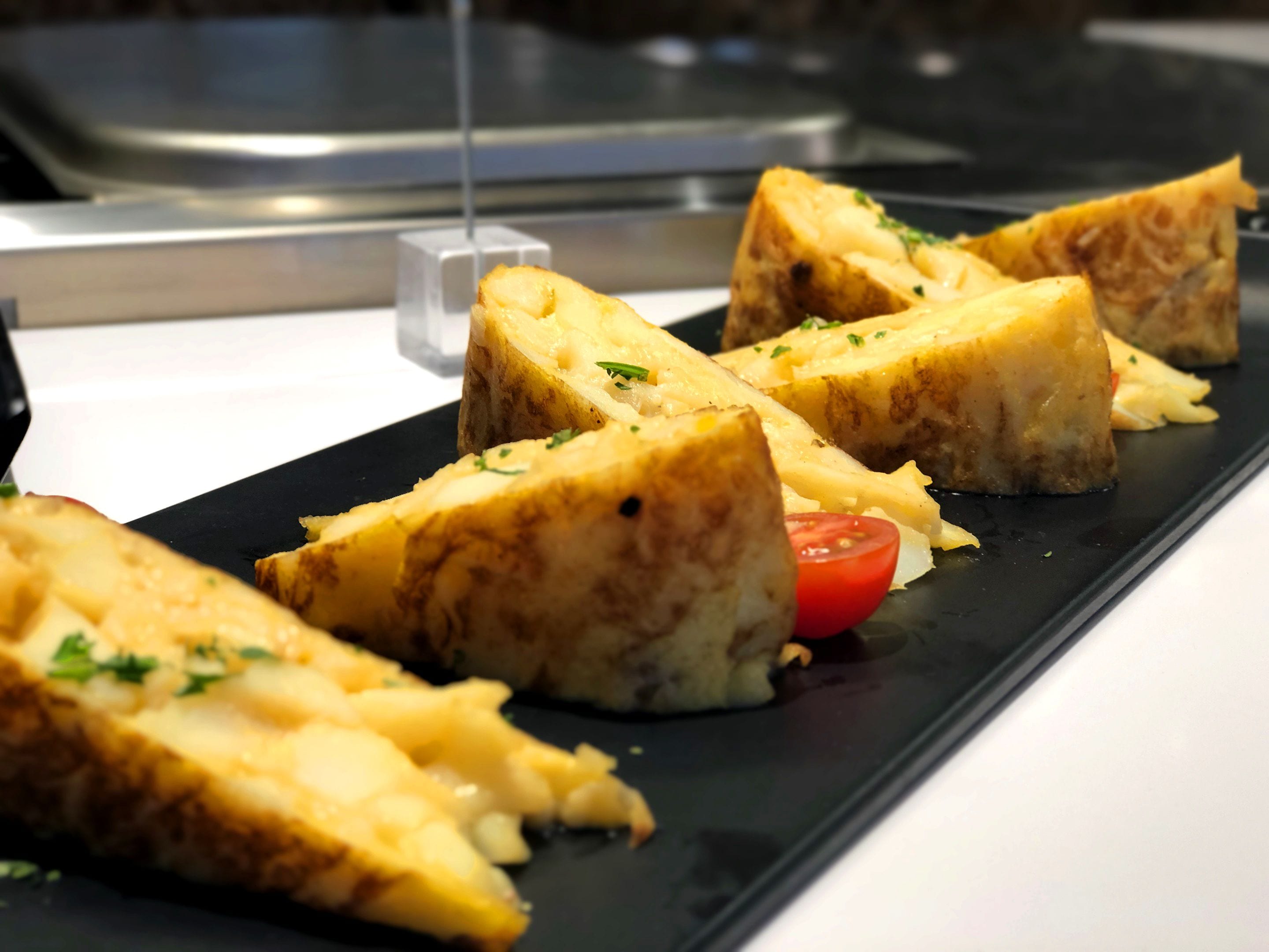 Um prato retangular preto com vários pedaços de comida assada dourada, guarnecido com ervas verdes e uma fatia de tomate ao lado.