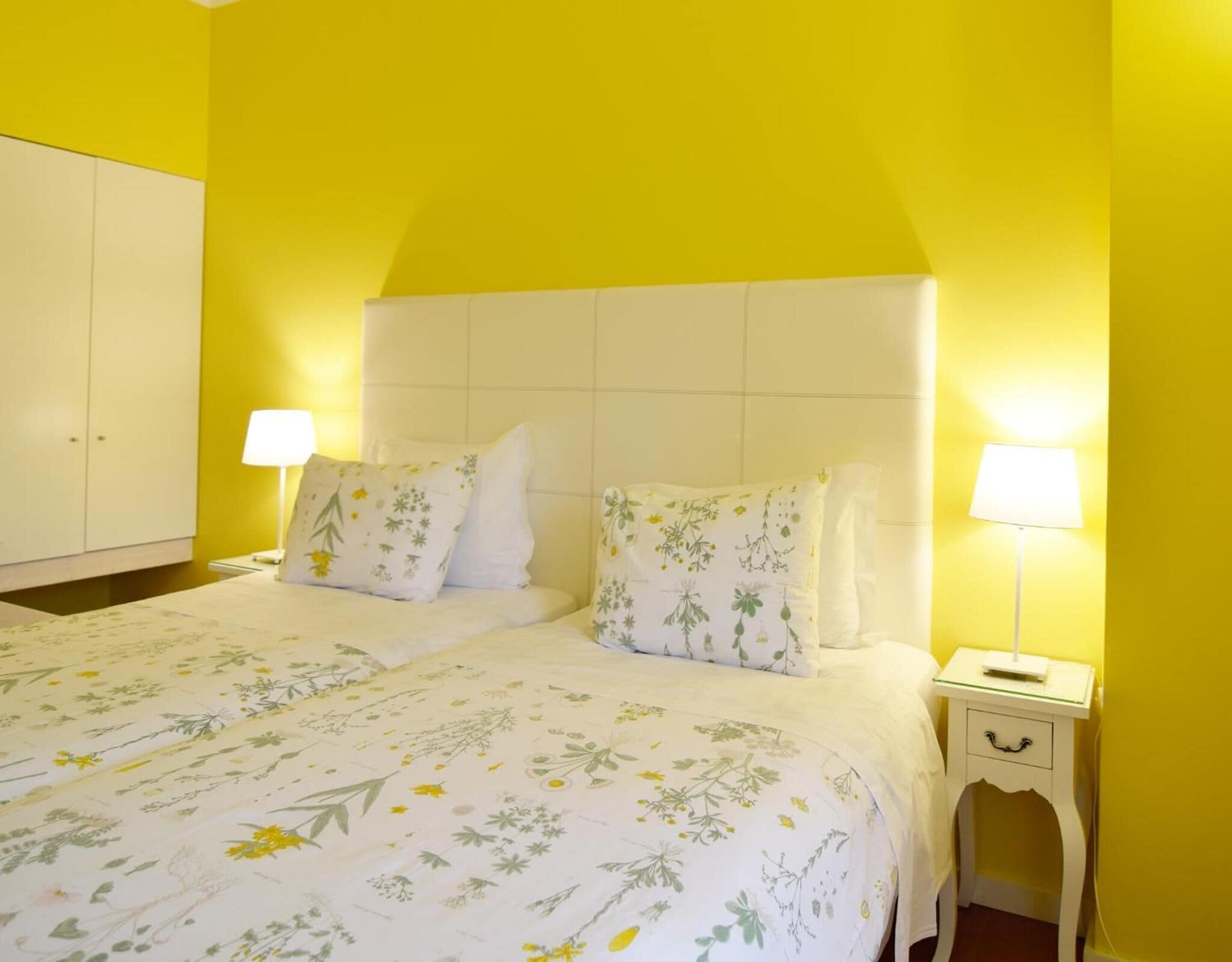 Cama de casal com roupa de cama floral, duas luminárias de cabeceira e um guarda-roupa branco num quarto com paredes amarelas.