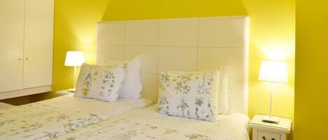 Cama de casal com roupa de cama floral, duas luminárias de cabeceira e um guarda-roupa branco num quarto com paredes amarelas.