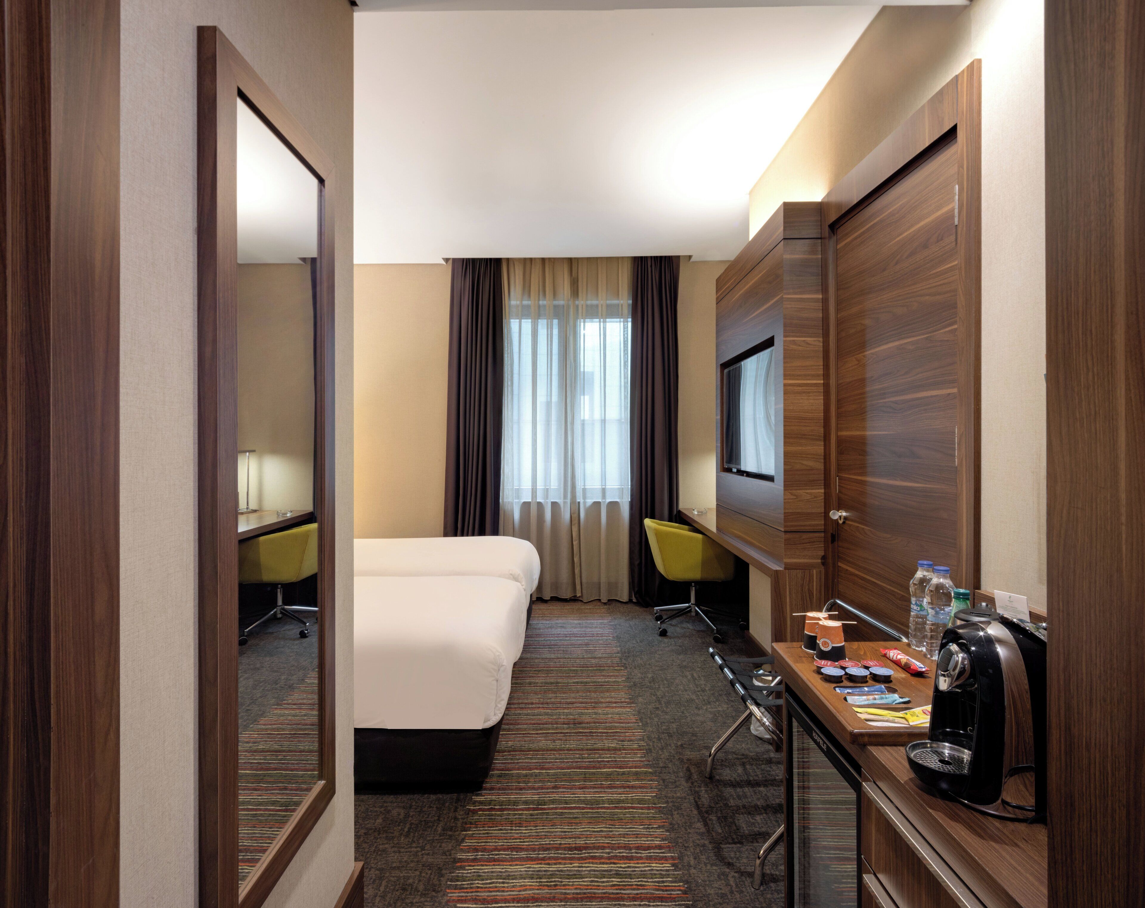 Quarto de hotel com uma cama, uma mesa com cafeteira, uma cadeira e uma televisão.