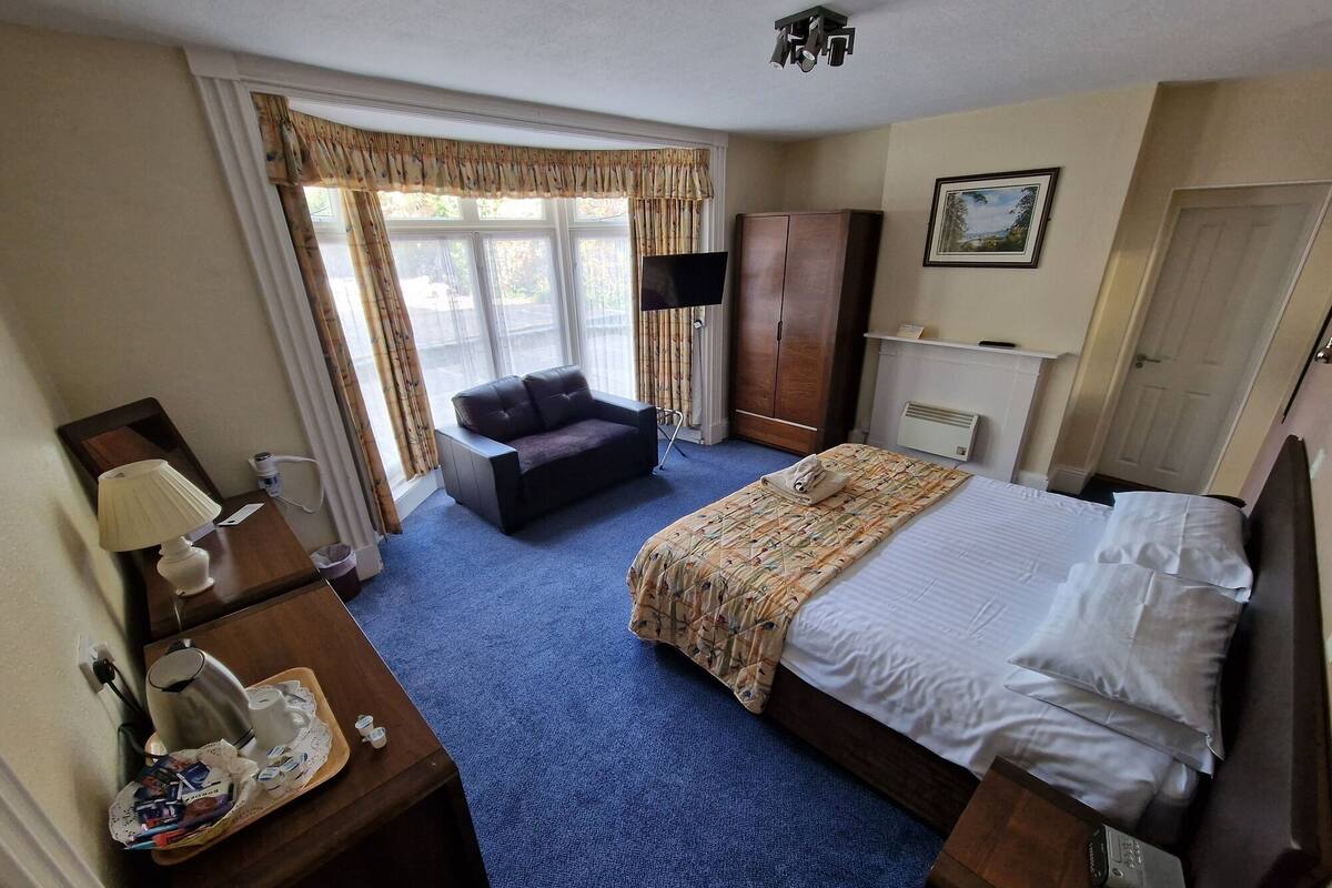 Quarto de hotel com uma cama grande, um sofá, uma mesa de trabalho com luminária, um guarda-roupa e uma janela com cortinas.