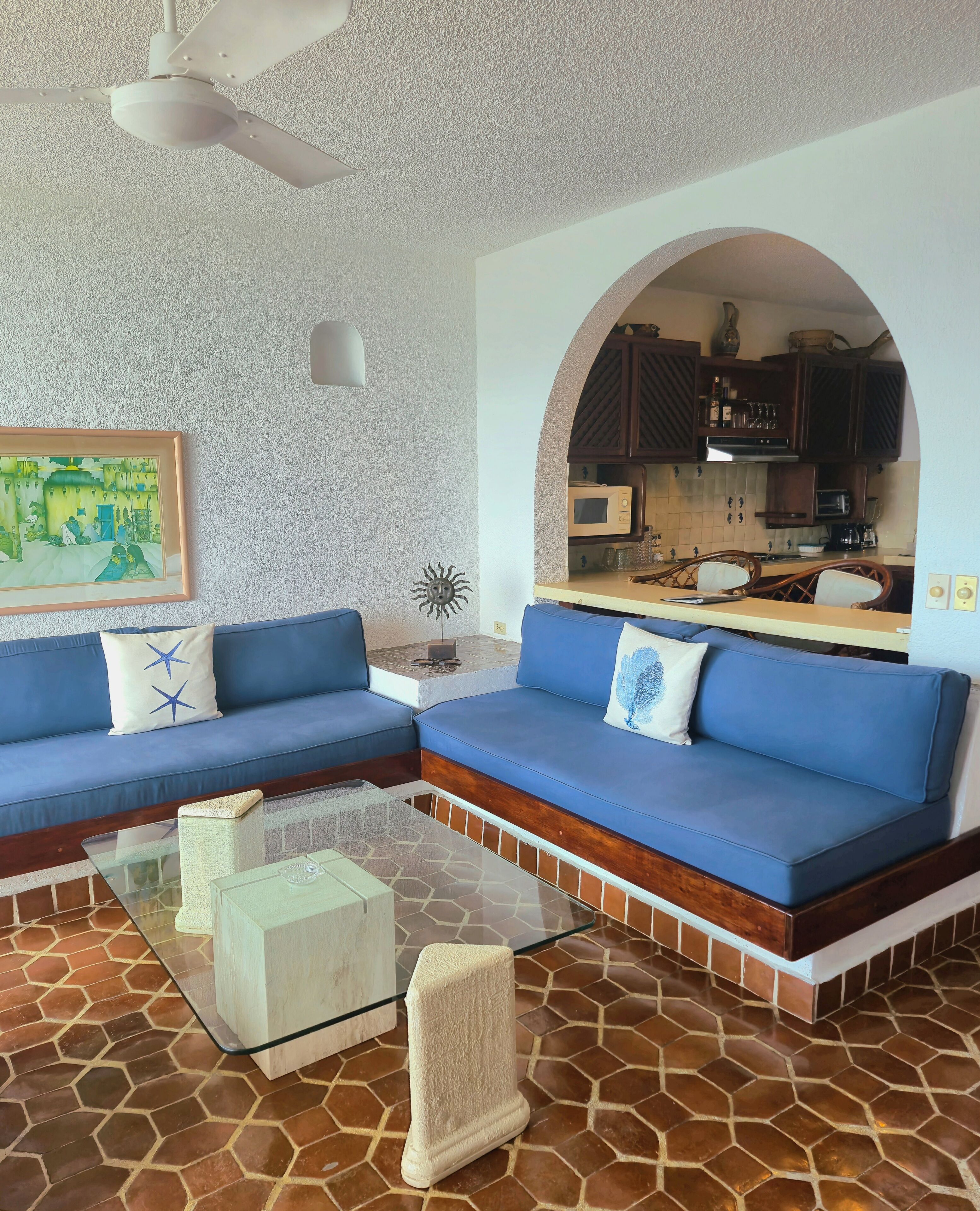 Sala de estar com sofá azul, mesa de centro de vidro e cozinha visível através de um arco.