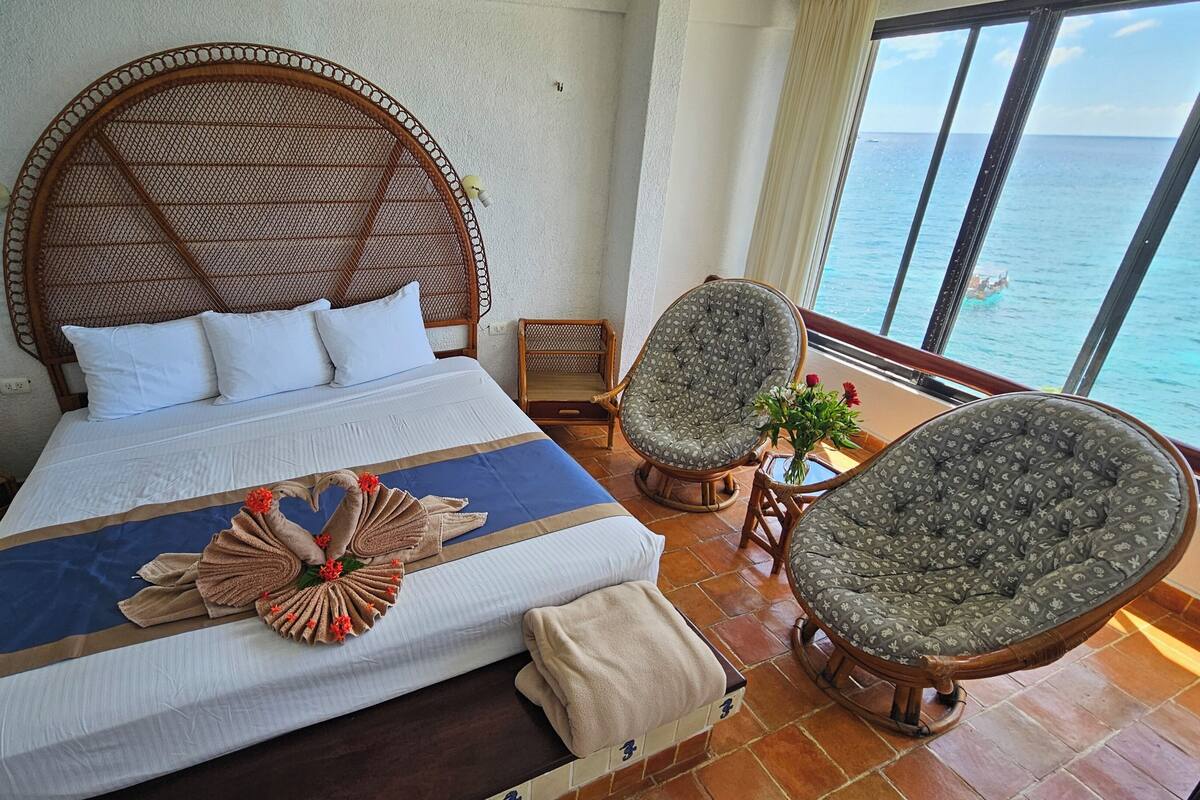 Quarto de hotel com uma cama grande, vista para o oceano, uma cadeira de vime e uma pequena mesa com um vaso de flores.