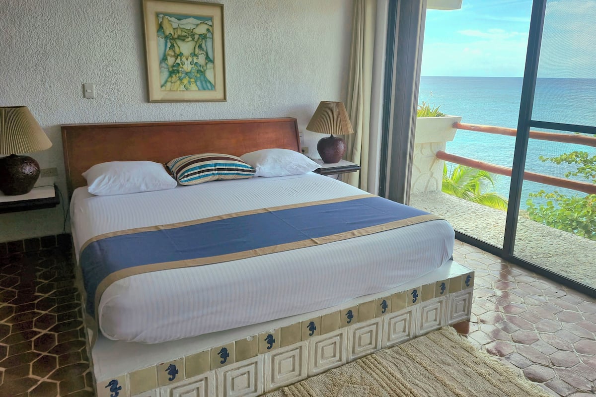 Quarto de hotel com uma cama grande, um quadro na parede, vista para o mar através de uma janela ampla e uma mesa de cabeceira com uma luminária.