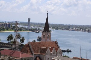Uma igreja costeira com uma torre alta, uma antena de rádio e vista para o mar e o horizonte urbano ao fundo.