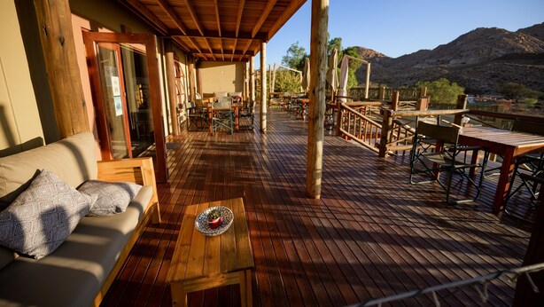 Um deck de madeira com mobiliário externo, incluindo um sofá e mesas, com vista para uma paisagem montanhosa.