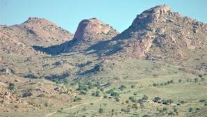 Paisagem acidentada com uma colina rochosa imponente e um caminho de terra que leva a um pequeno povoado.
