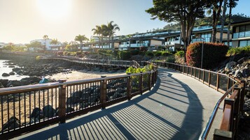 Uma passarela de madeira que leva a uma praia com costa rochosa, palmeiras e edifícios modernos ao fundo.