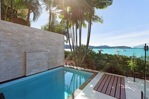Piscina com muro de pedra, deck de madeira e plantas tropicais.