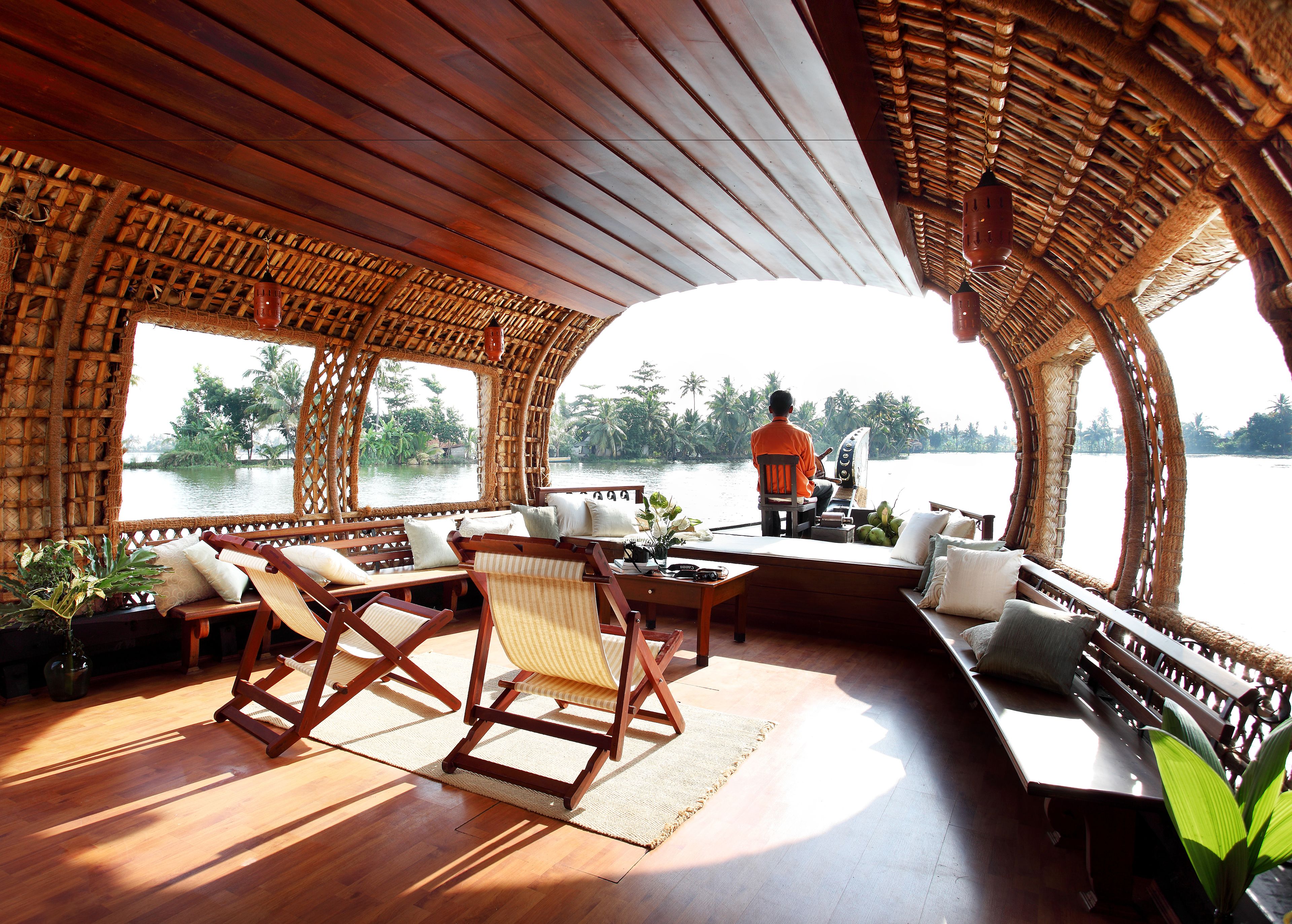 Interior de barco com mobiliário de madeira, área de estar e vista para um rio com palmeiras.