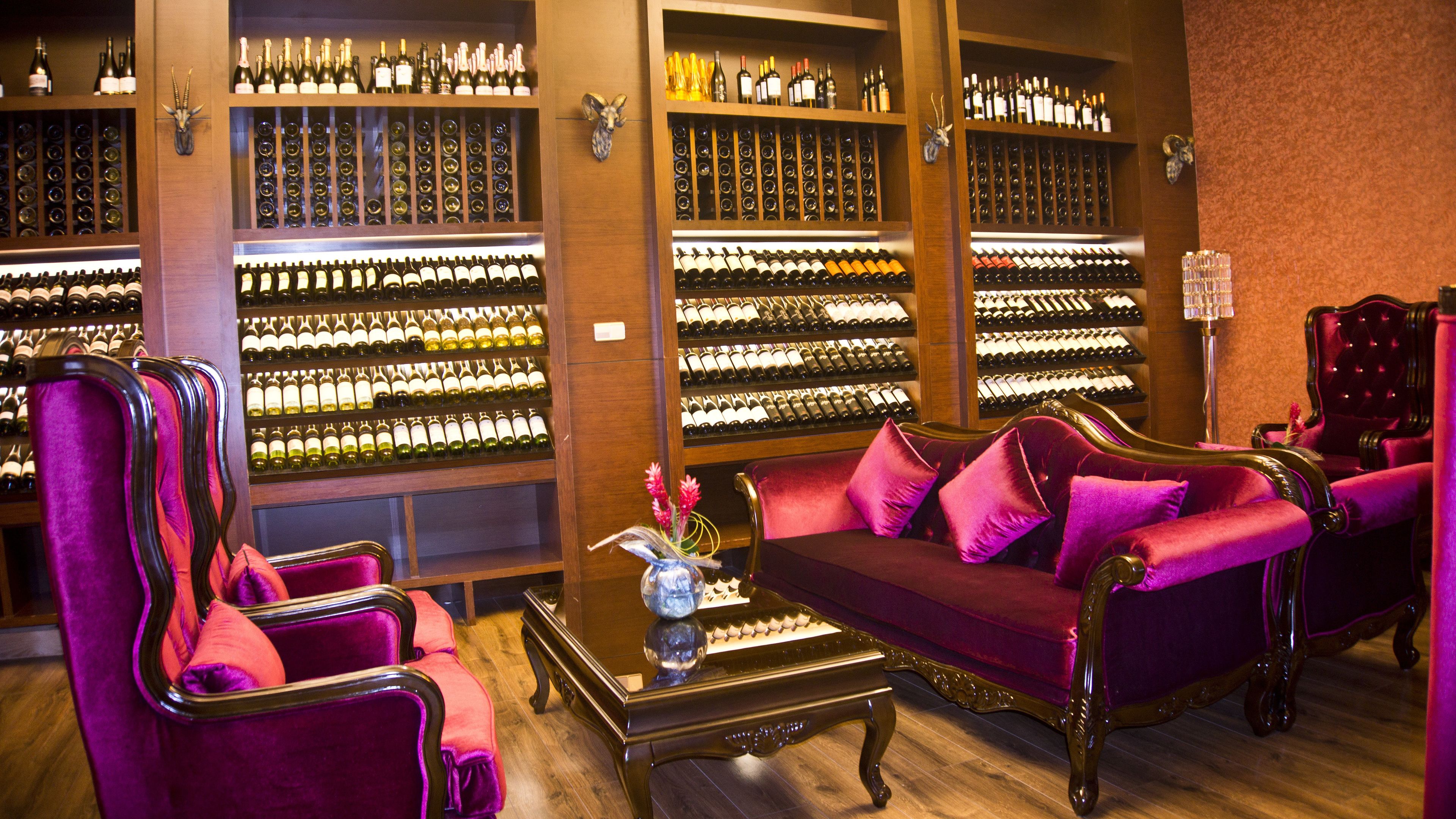 Um quarto luxuoso com um sofá de veludo vermelho, poltronas a jogo e uma estante de madeira cheia de garrafas de vinho.