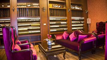 Um quarto luxuoso com um sofá de veludo vermelho, poltronas a jogo e uma estante de madeira cheia de garrafas de vinho.