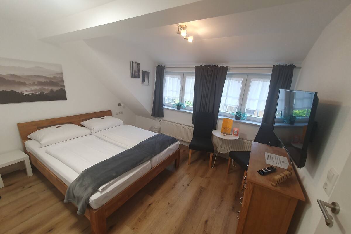 Quarto com cama, escrivaninha, cadeira, televisão e janela com cortinas.