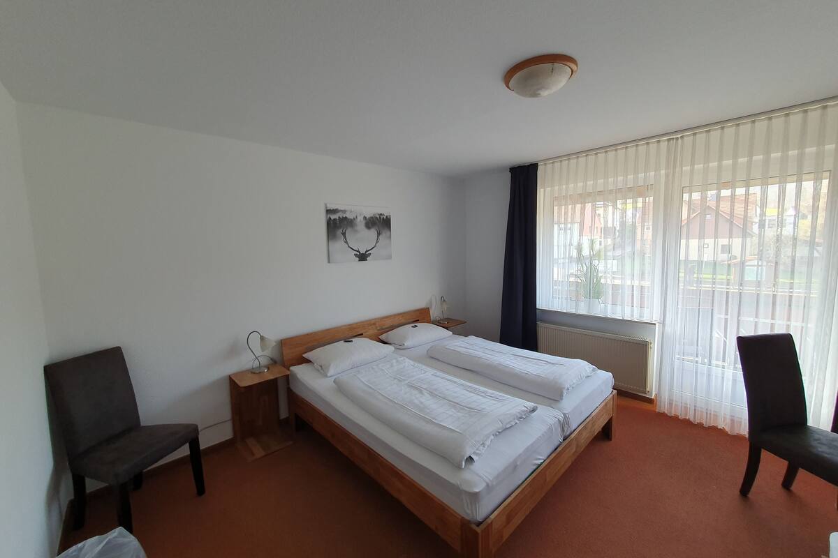 Quarto de hotel com uma cama de madeira, uma mesa de cabeceira, uma cadeira, uma janela com cortinas leves e uma obra de arte pendurada na parede.