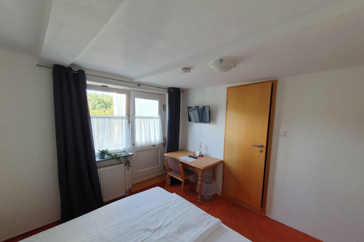 Quarto com cama, escrivaninha, televisão e janela com cortinas.