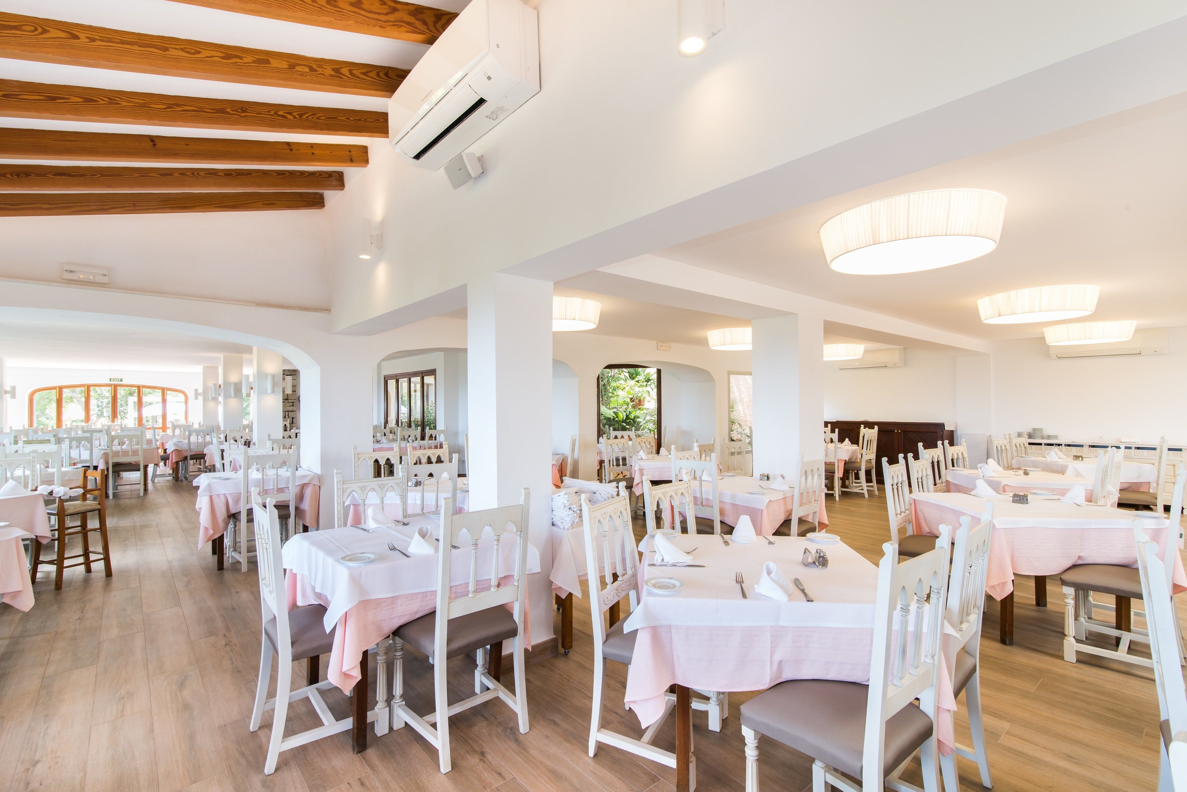 Uma sala de jantar espaçosa, com mesas e cadeiras brancas, vigas de madeira no teto e luz natural proveniente de grandes janelas.