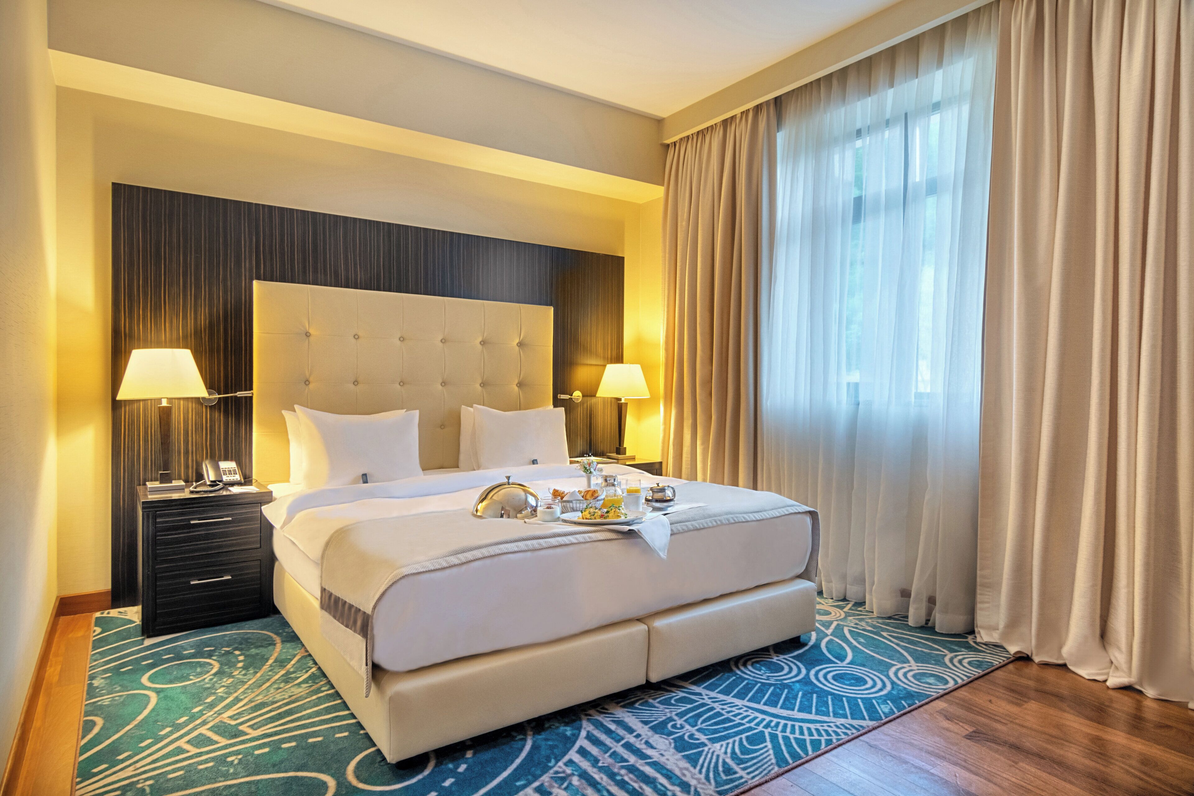 Quarto de hotel com uma cama grande, duas luminárias de cabeceira, mesa de cabeceira e janela com cortinas.
