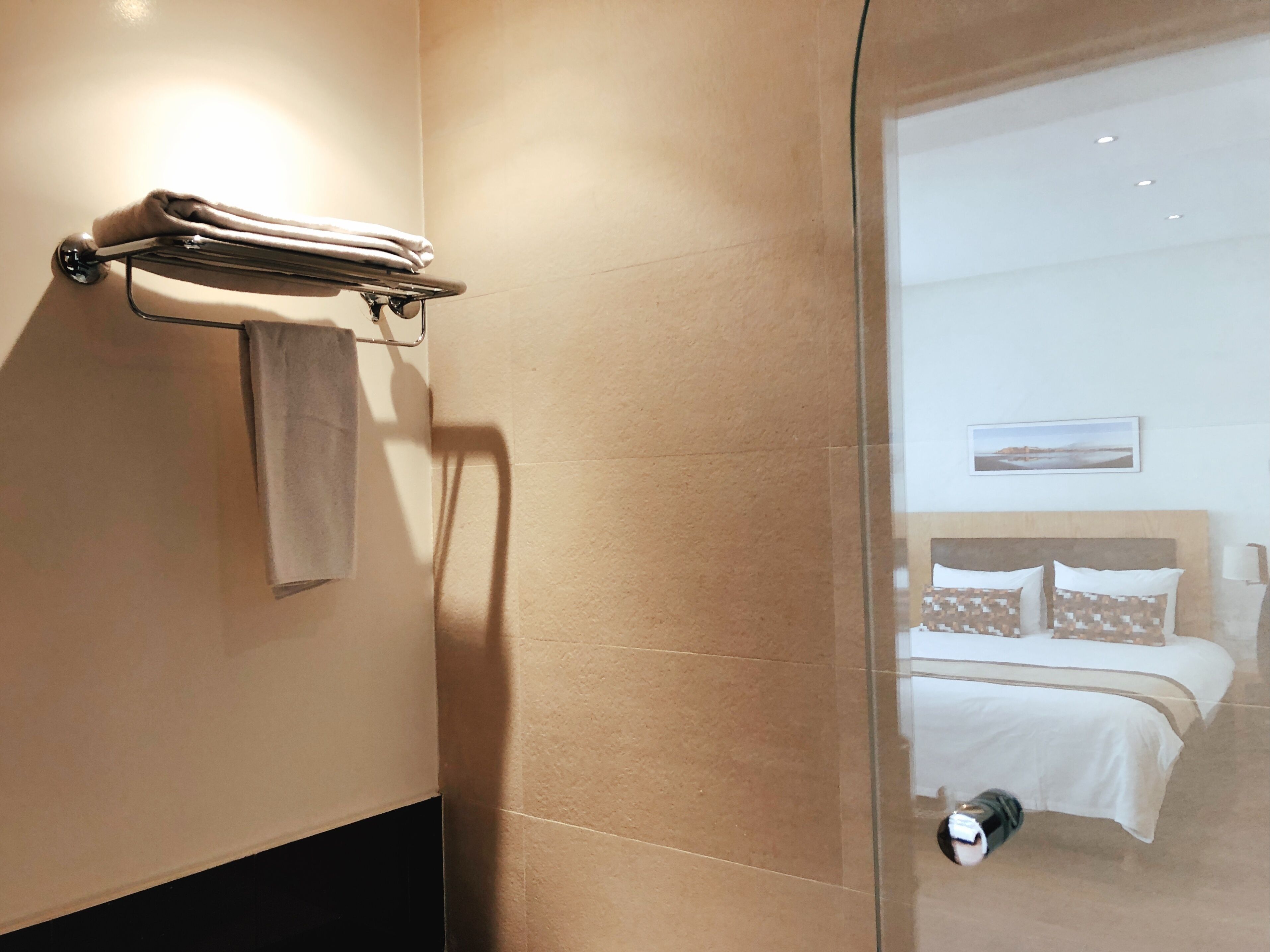 Casa de banho de hotel com cabine de chuveiro de vidro, porta-toalhas com toalhas dobradas e quarto visível com cama e mesa de cabeceira.