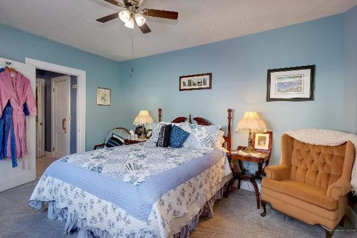 Um quarto com uma cama, uma cadeira, uma mesinha de cabeceira e um ventilador de teto.