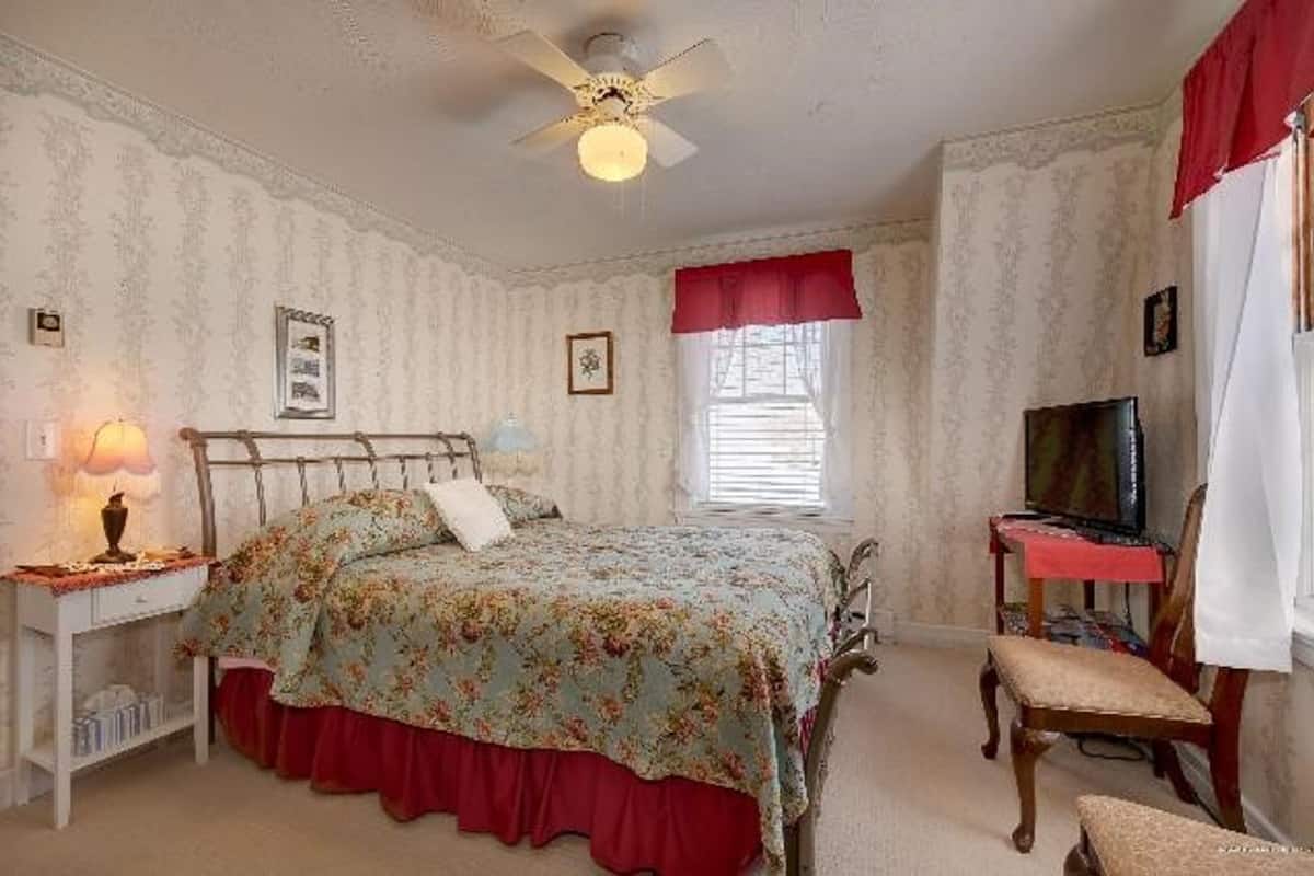 Quarto com cama, escrivaninha, cadeira, televisão e ventoinha no teto.