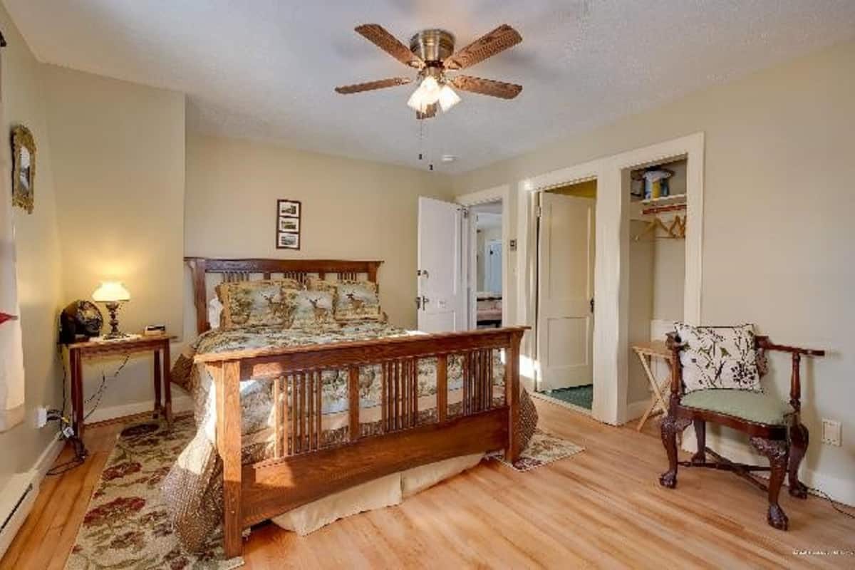 Quarto com cama de madeira, ventilador de teto, mesa de cabeceira, abajur, cadeira e porta que dá para outro cômodo.