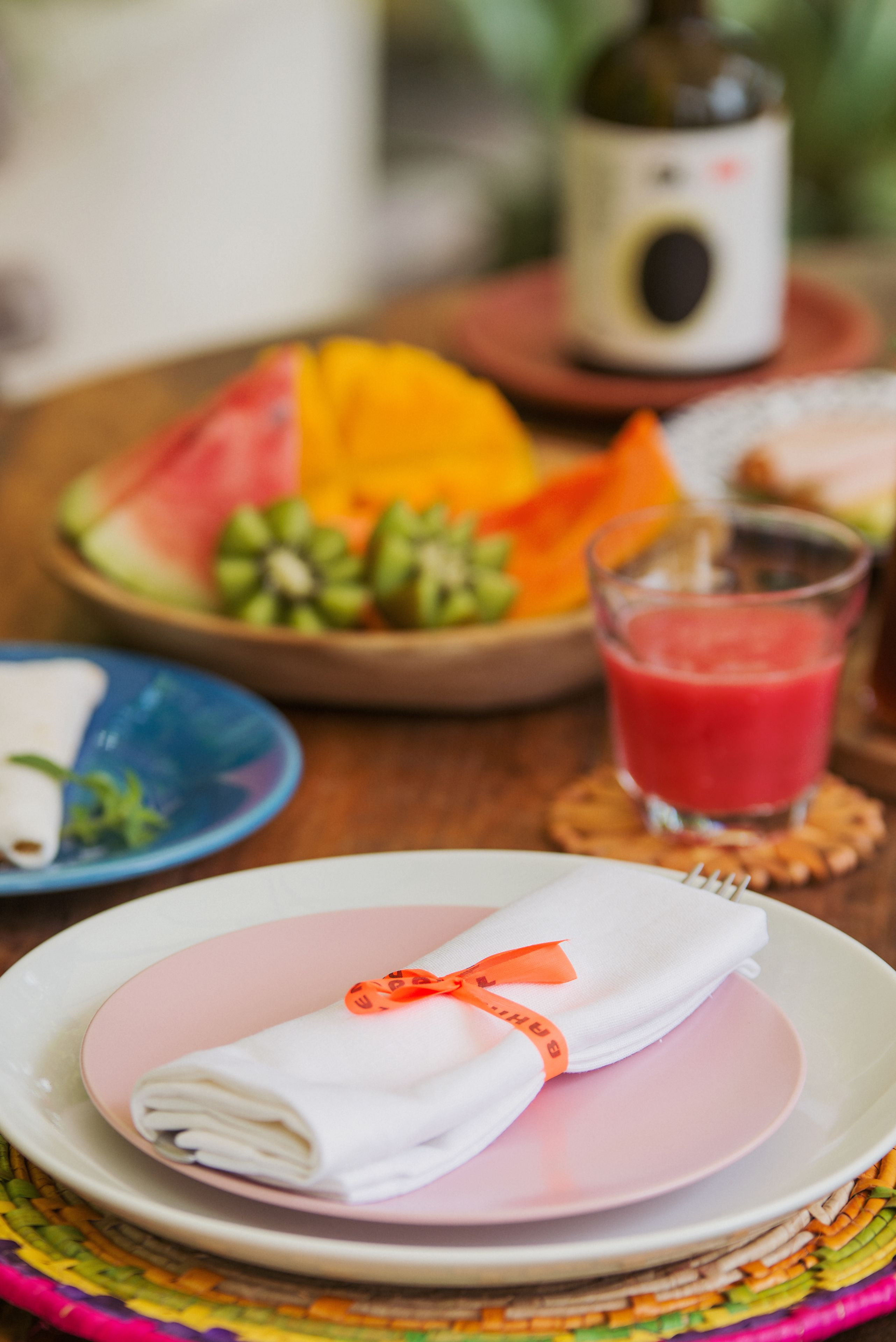 Uma mesa com uma toalha de papel branca dobrada sobre um prato rosa, um copo de suco vermelho e um prato com frutas fatiadas.