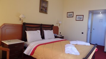 Quarto de hotel com cabeceira de madeira, duas luminárias de apoio, cama com roupa de cama branca e manta vermelha, e uma porta.