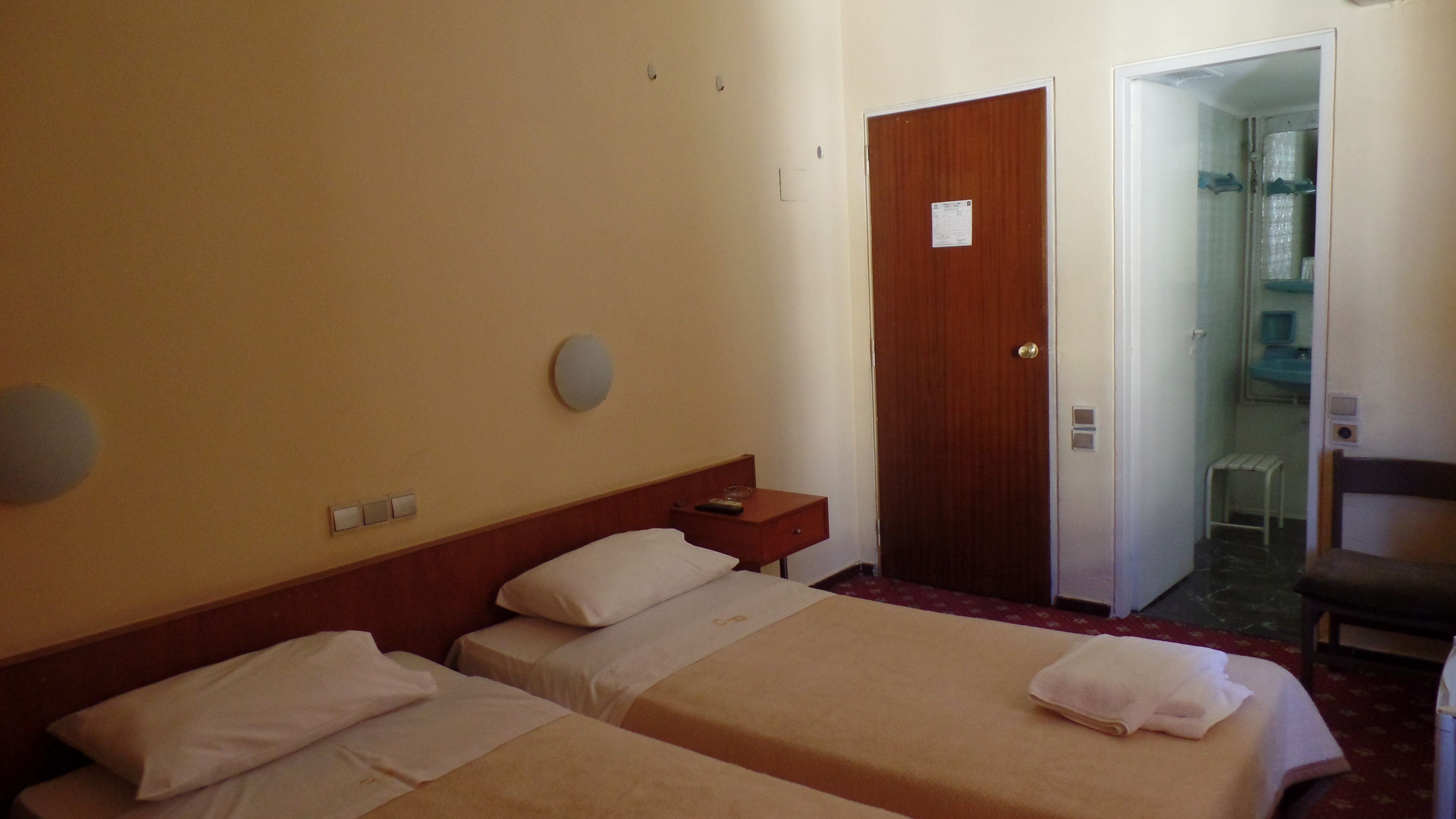 Quarto de hotel com duas camas, uma mesa de cabeceira de madeira, uma luminária fixada na parede e uma porta que dá para o banheiro.